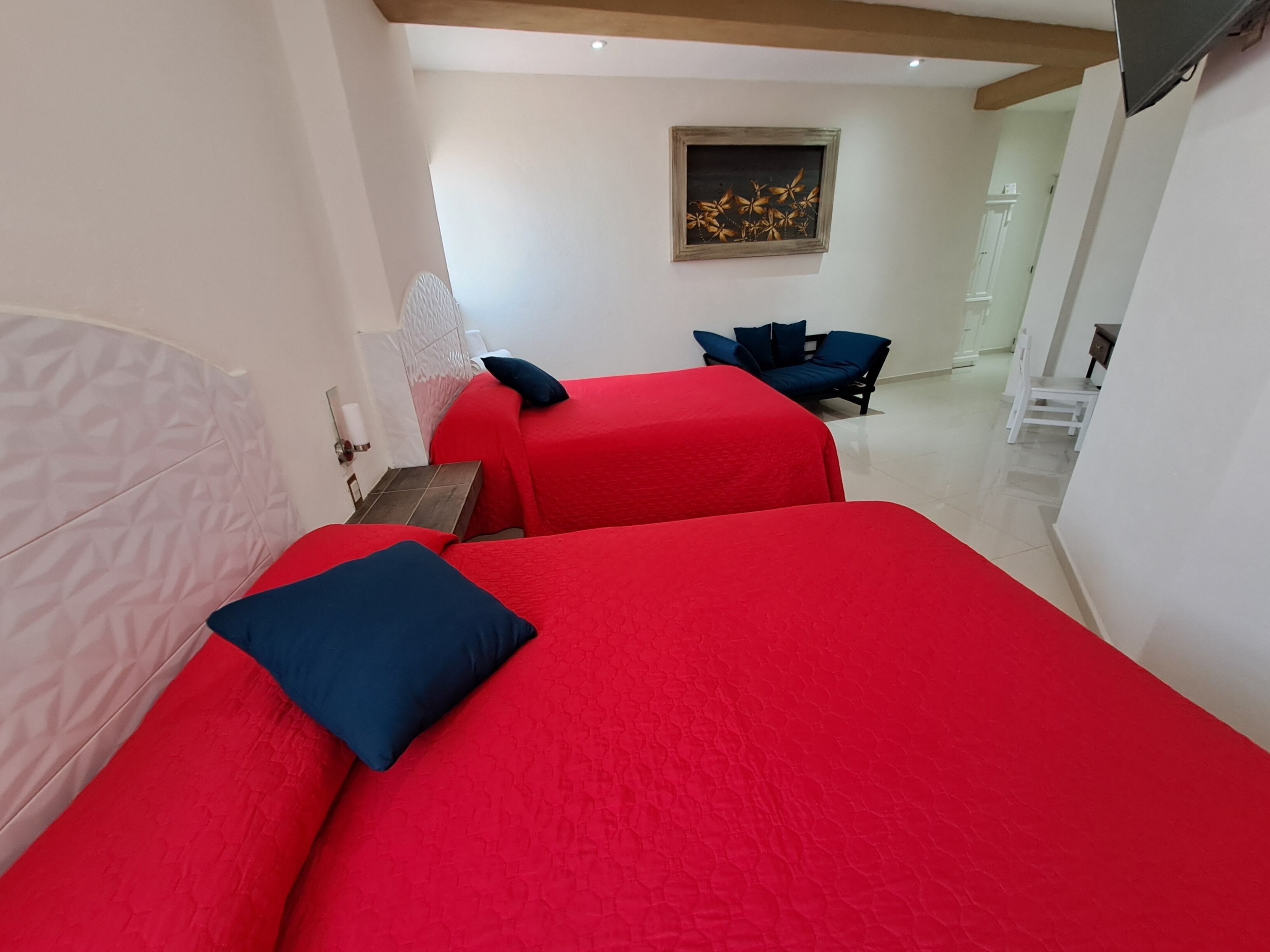 Quarto de hotel com duas camas vermelhas, uma cadeira azul e um quadro na parede.
