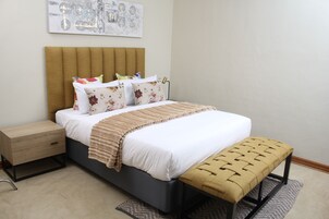 Quarto com cama, mesa de cabeceira, banco e uma obra de arte na parede.