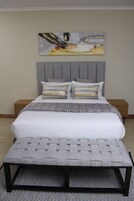 Um quarto bem arrumado com uma cama grande, um banco estofado cinza e uma pintura abstrata moderna na parede.