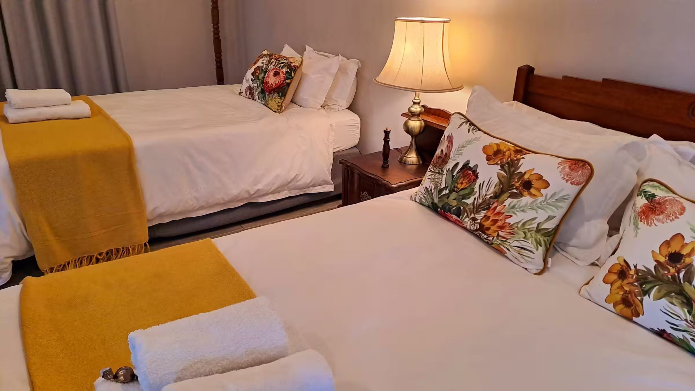 Quarto de hotel com duas camas, cada uma com roupa de cama branca e almofadas estampadas com flores. Há uma mesa de cabeceira com uma luminária e um pequeno objeto decorativo.
