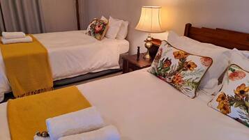 Quarto de hotel com duas camas, cada uma com roupa de cama branca e almofadas estampadas com flores. Há uma mesa de cabeceira com uma luminária e um pequeno objeto decorativo.
