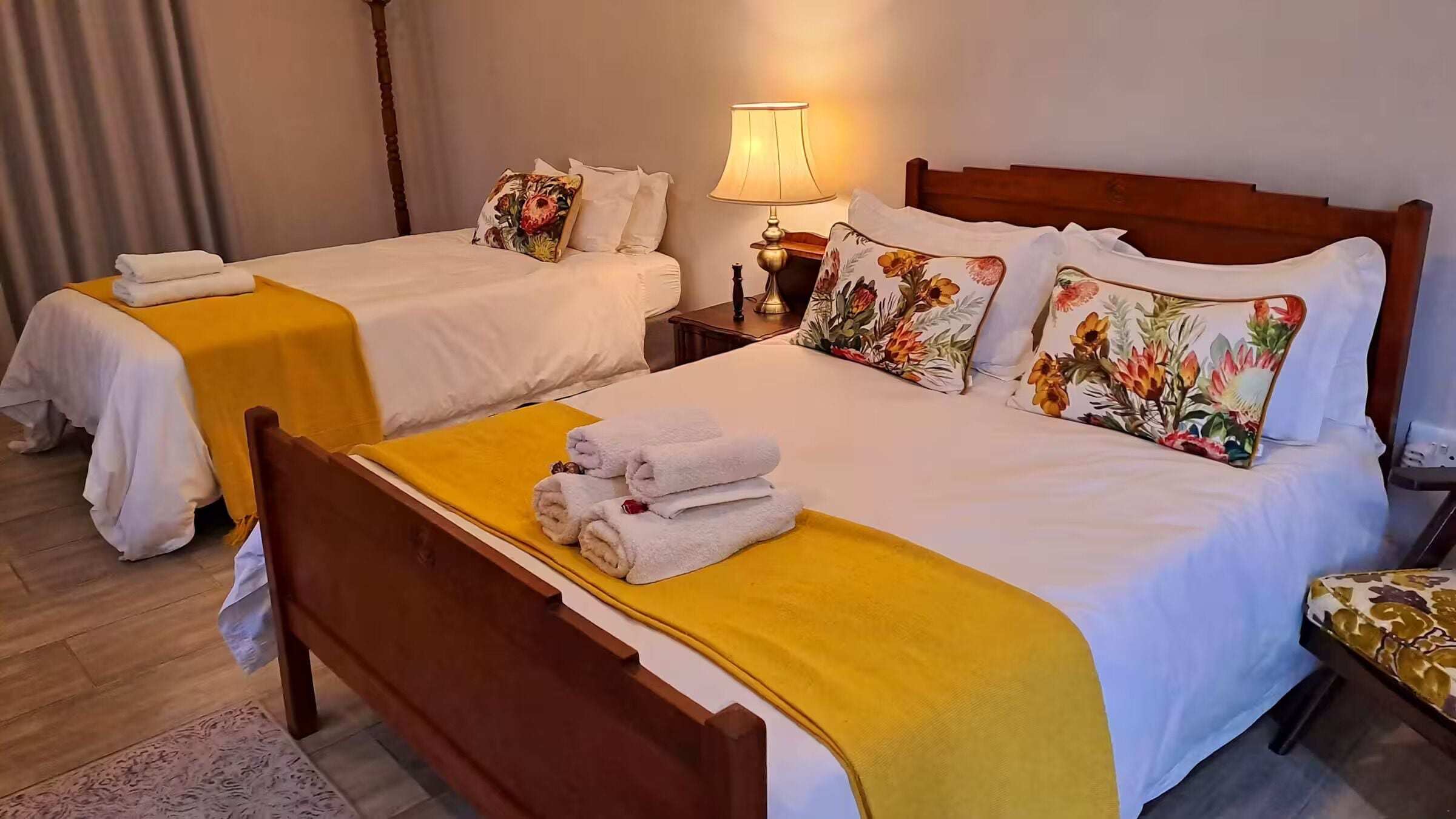 Quarto de hotel com duas camas, cada uma com roupa de cama branca e almofadas com padrão floral. Há uma mesa de cabeceira com abajur e uma pequena mesa com artigos de higiene pessoal.
