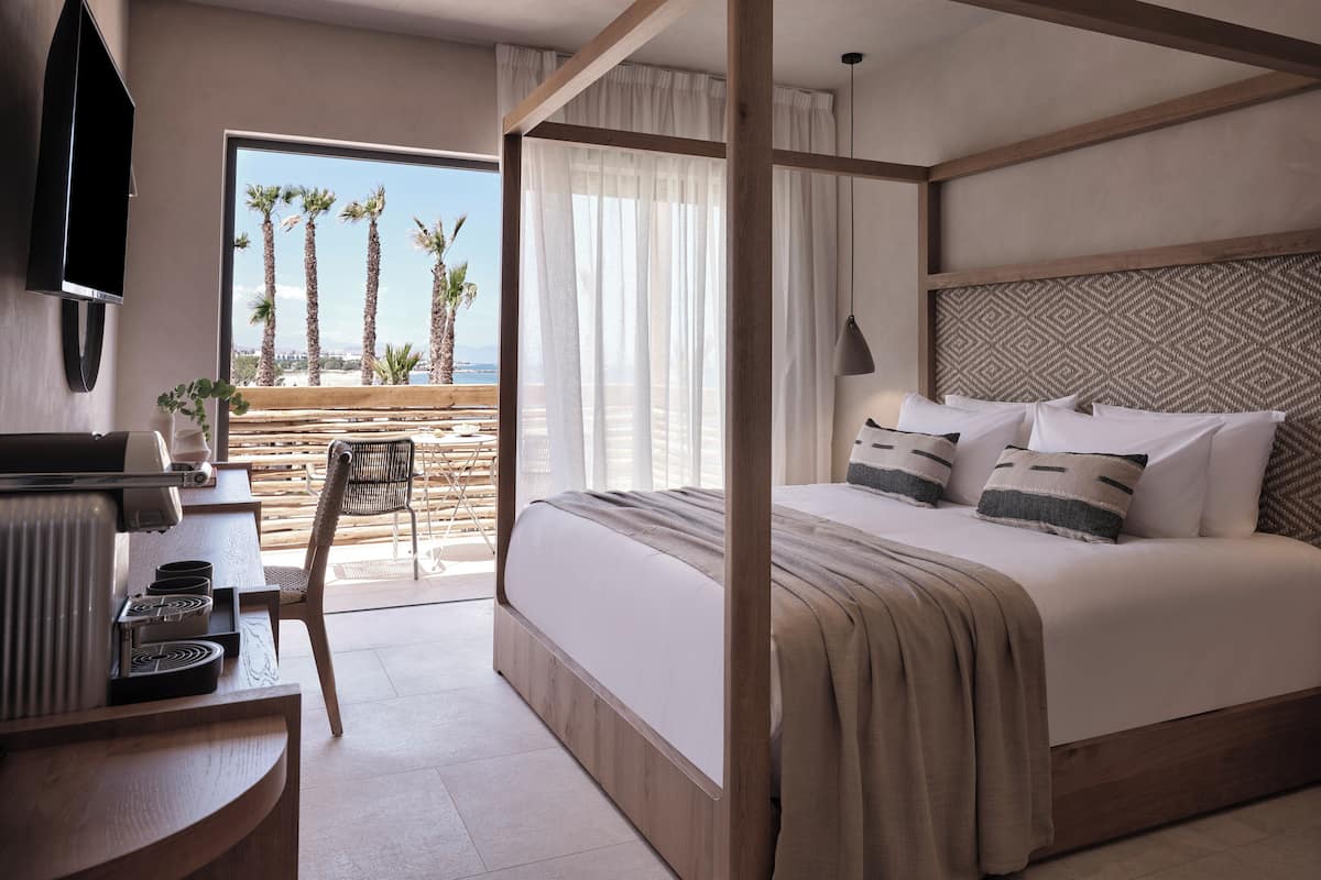 Quarto de hotel moderno com uma cama grande, uma mesa com televisão de tela plana e uma varanda com vista para palmeiras e a praia.