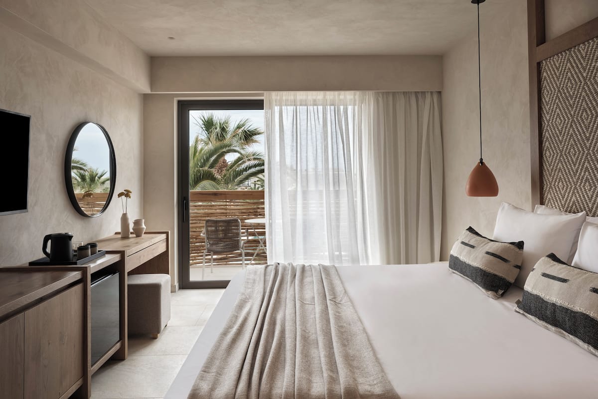 Quarto de hotel moderno com uma cama grande, uma escrivaninha com cafeteira, televisão e varanda com vista para palmeiras.