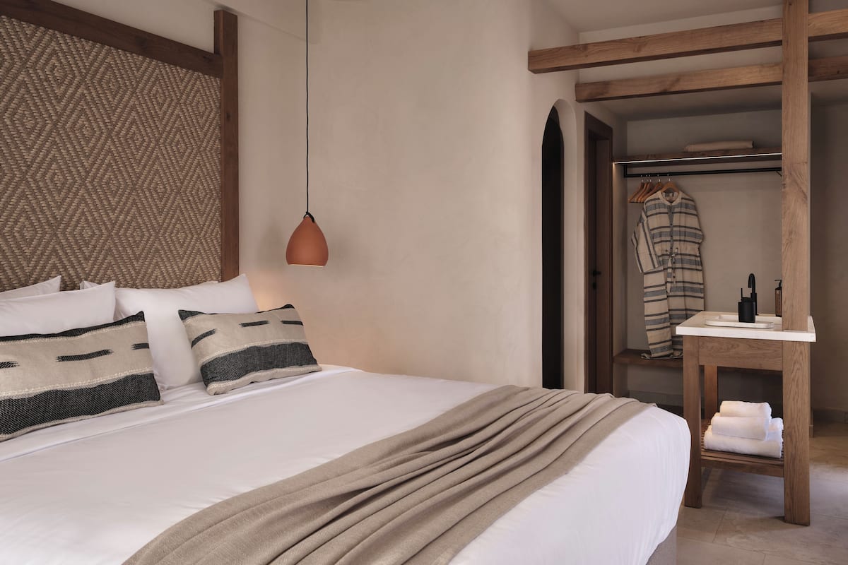 Cama bem arrumada com cabeceira estampada, luminária pendente e um banco de madeira com toalhas.