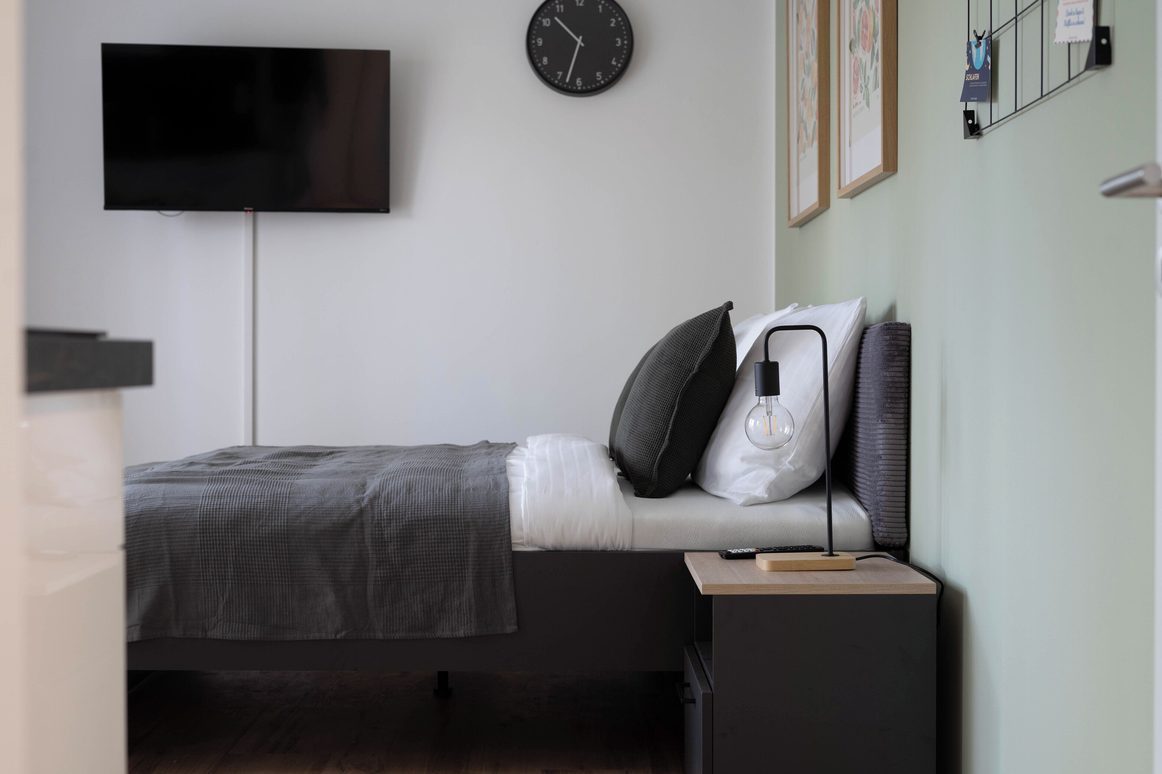 Quarto moderno com cama, mesa de cabeceira, luminária e TV fixada na parede.