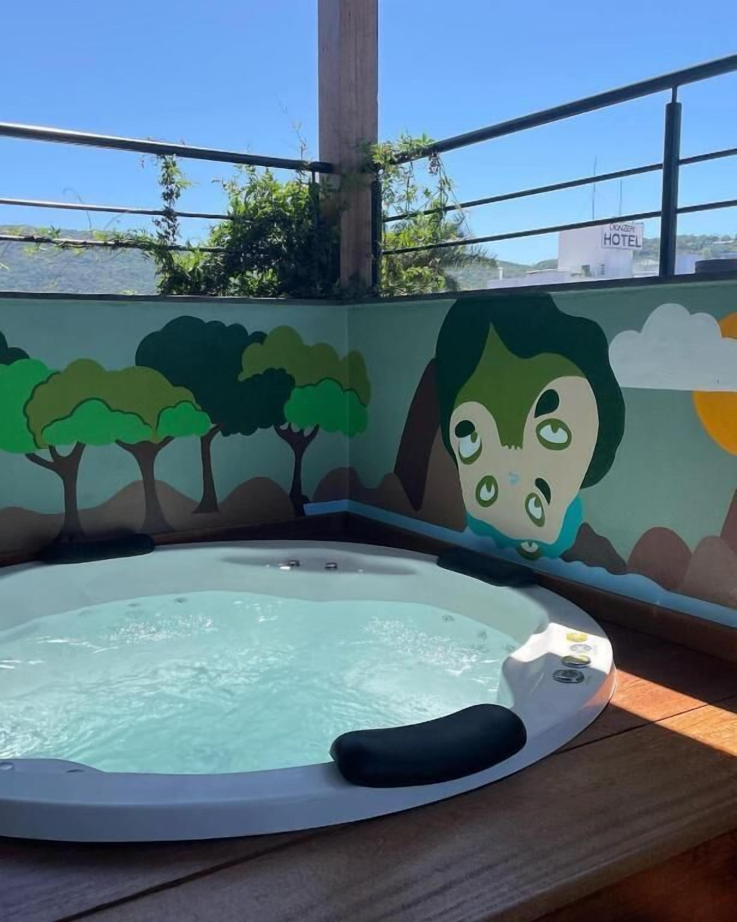 Uma banheira de hidromassagem com um mural de árvores e um rosto, cercada por um deck de madeira e guarda-corpo de metal.