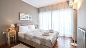 Quarto com cama, mesas de cabeceira, luminária, radiador e janela com cortinas.