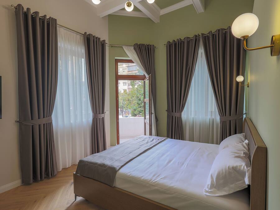 Quarto com cama, cortinas, luminária na parede e vista pela janela.