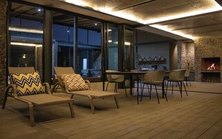 Uma área externa com assentos de lounge e uma mesa, uma lareira de pedra e amplas janelas com vista para um edifício.