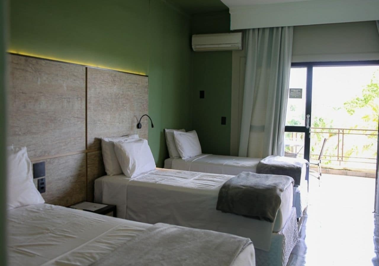 Quarto de hotel com três camas, uma janela grande com cortinas e um aparelho de ar condicionado na parede.