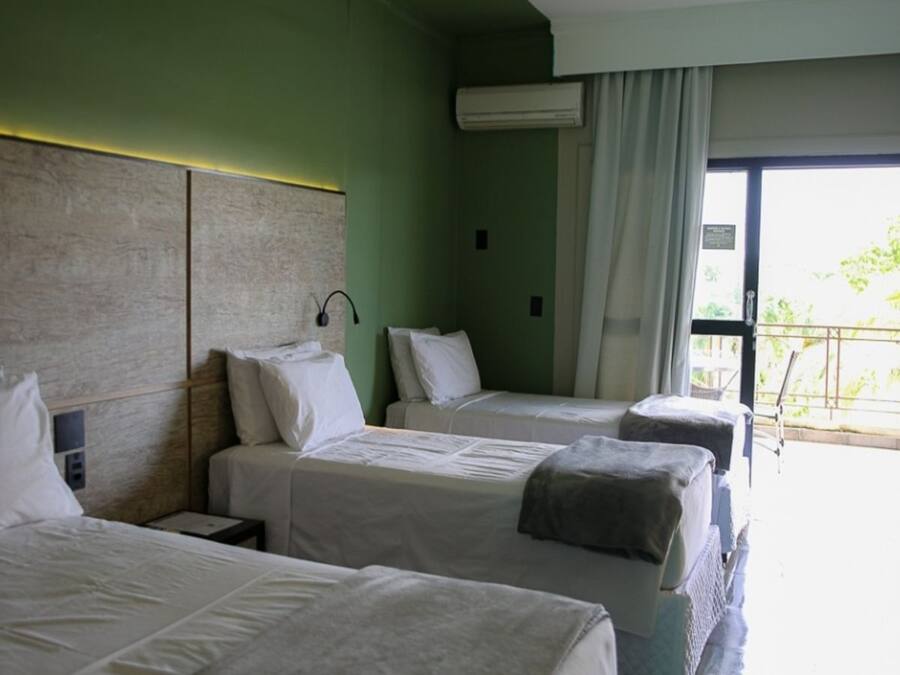 Quarto de hotel com três camas, uma janela grande com cortinas e um aparelho de ar condicionado na parede.