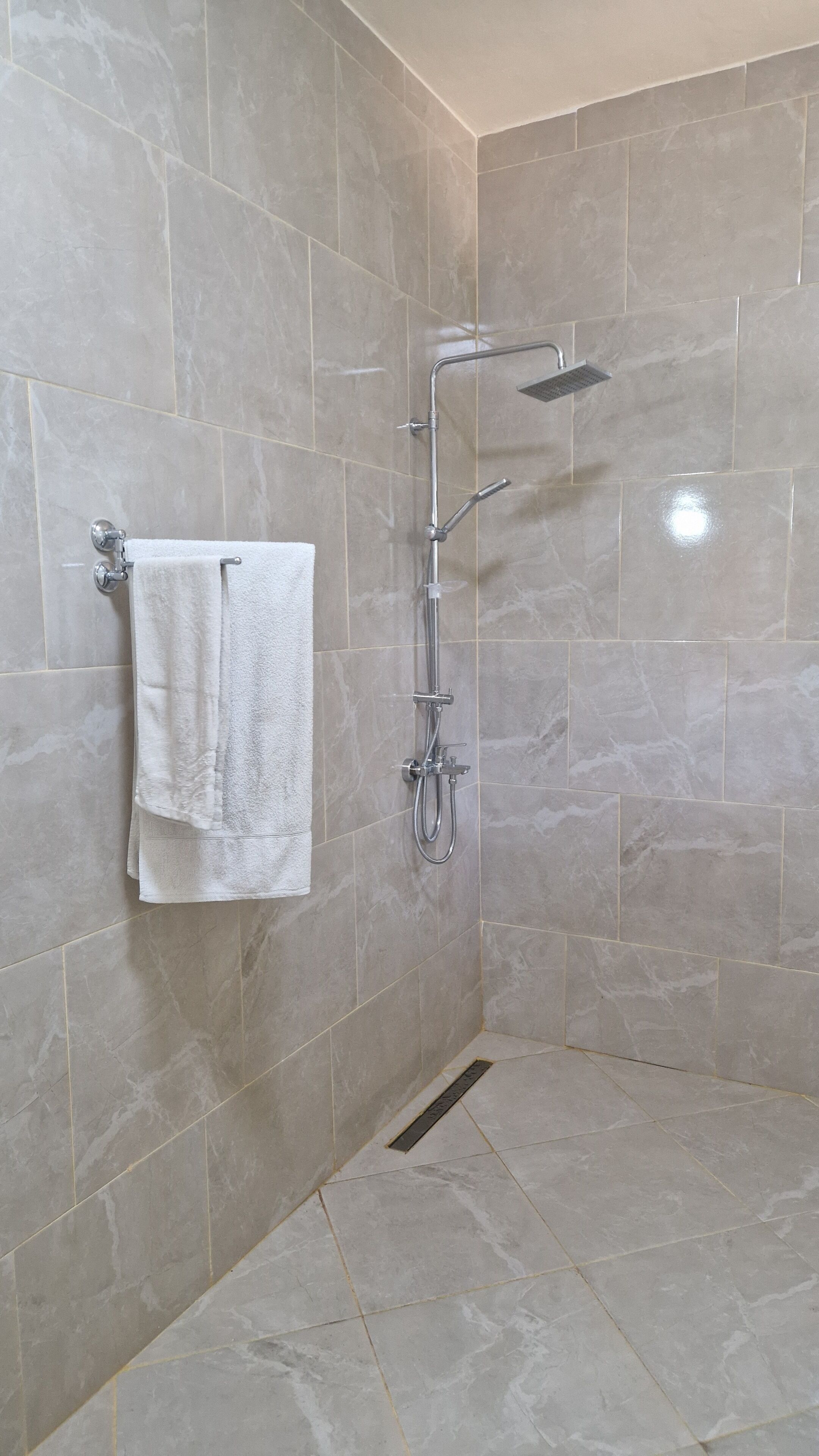 Área de chuveiro com chuveiro manual, porta-toalhas fixado na parede e uma toalha branca, e ralo no chão.