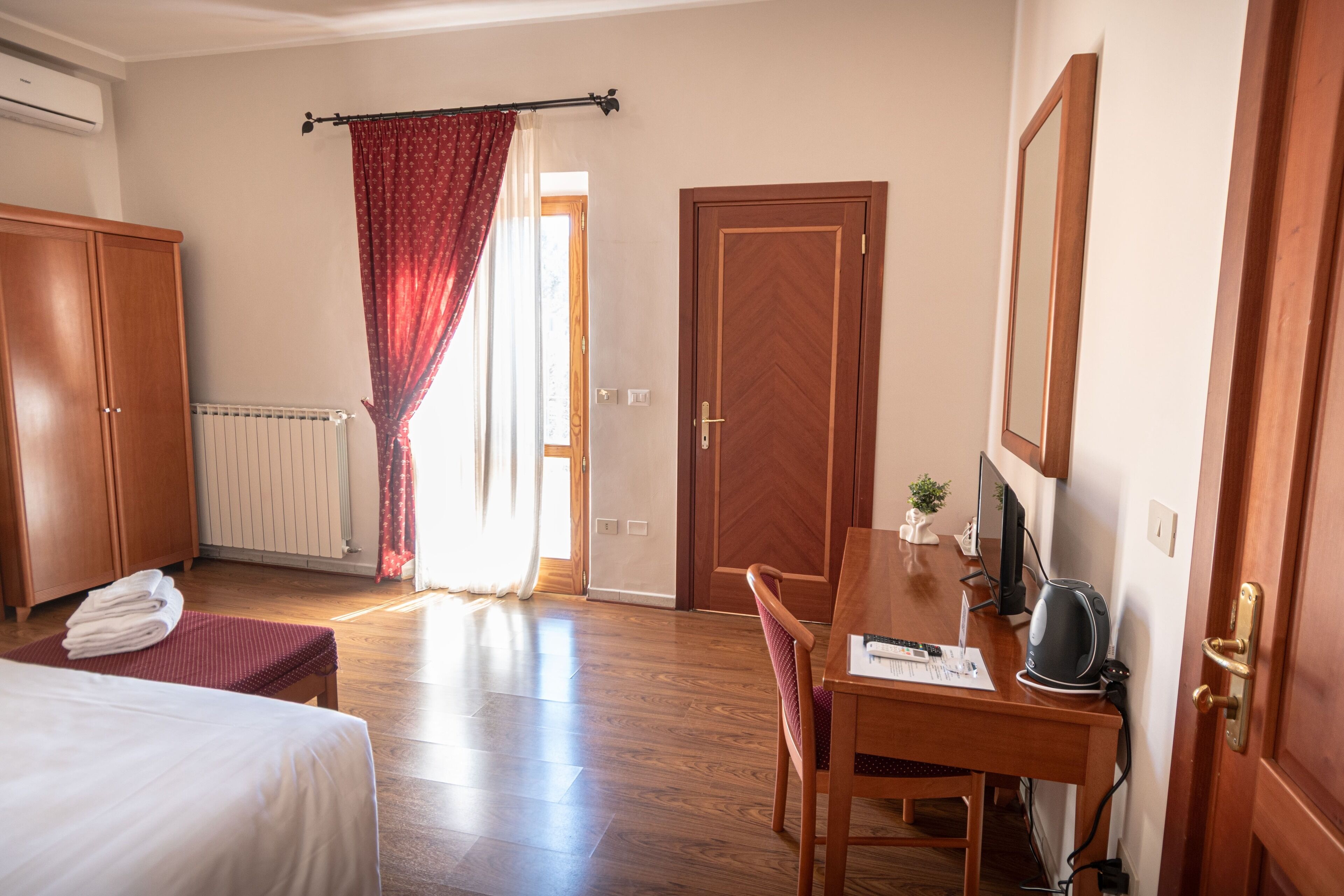 Quarto de hotel com cama, escrivaninha com computador e chaleira, armário e janela com cortinas.