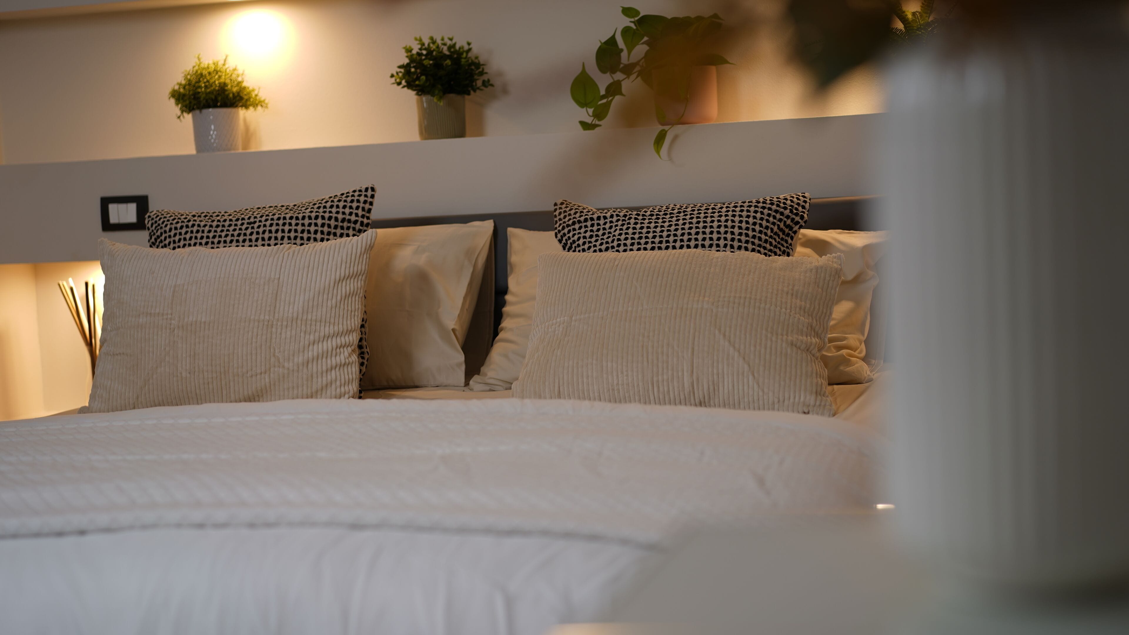 Uma cama bem arrumada com edredom branco e vários travesseiros, incluindo um com padrão.
