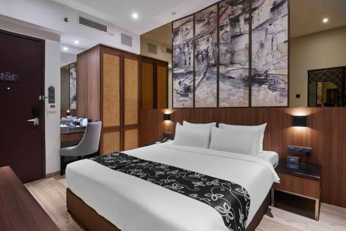 Quarto de hotel com uma cama grande, uma escrivaninha com cadeira, um guarda-roupa e uma obra de arte pendurada na parede.