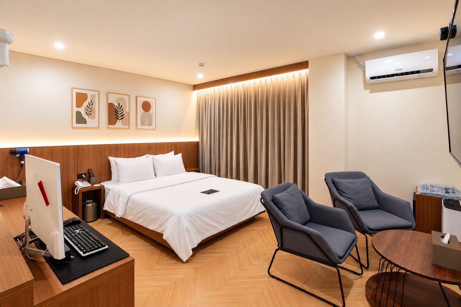 Quarto de hotel com cama, escrivaninha, computador, cadeira e ar condicionado.