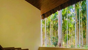 Quarto com um banco de madeira, uma mesa pequena e uma janela com vista para palmeiras altas.