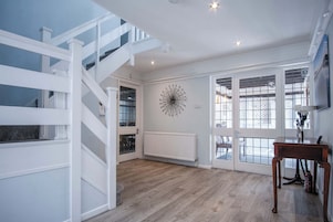 Quarto amplo com piso de madeira, escada branca, radiador e uma janela grande com um detalhe decorativo na parede.