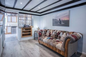 Uma cozinha moderna com armários de madeira e forno embutido, uma sala de estar aconchegante com um sofá grande e estofado, e vista para o mar através de uma janela ampla.