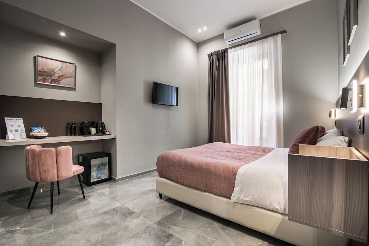 Quarto de hotel moderno com cama, escrivaninha com computador, cadeira, televisão e ar condicionado.