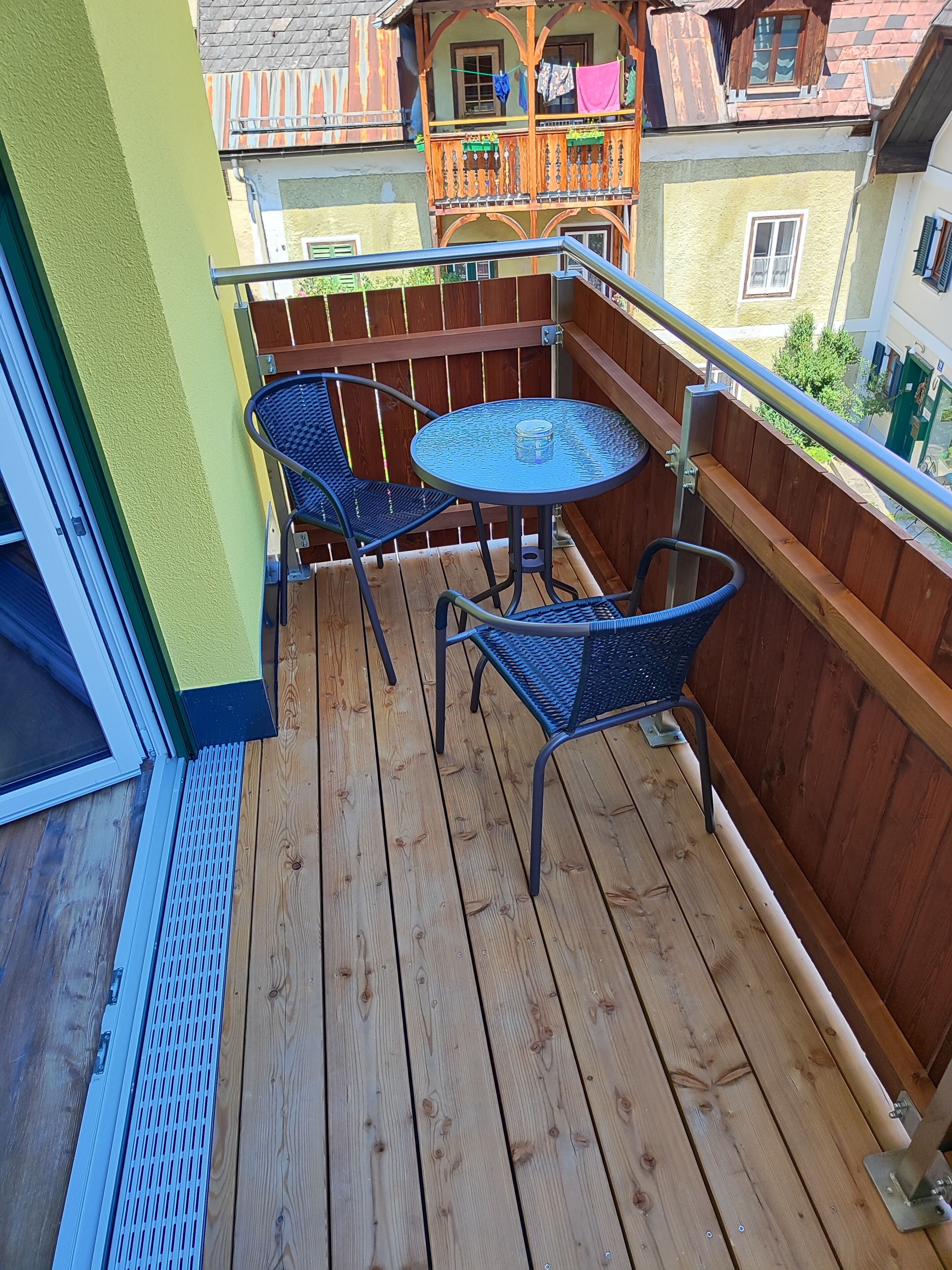 Um deck de madeira com duas cadeiras e uma pequena mesa, uma cerca de madeira e vista para edifícios próximos.