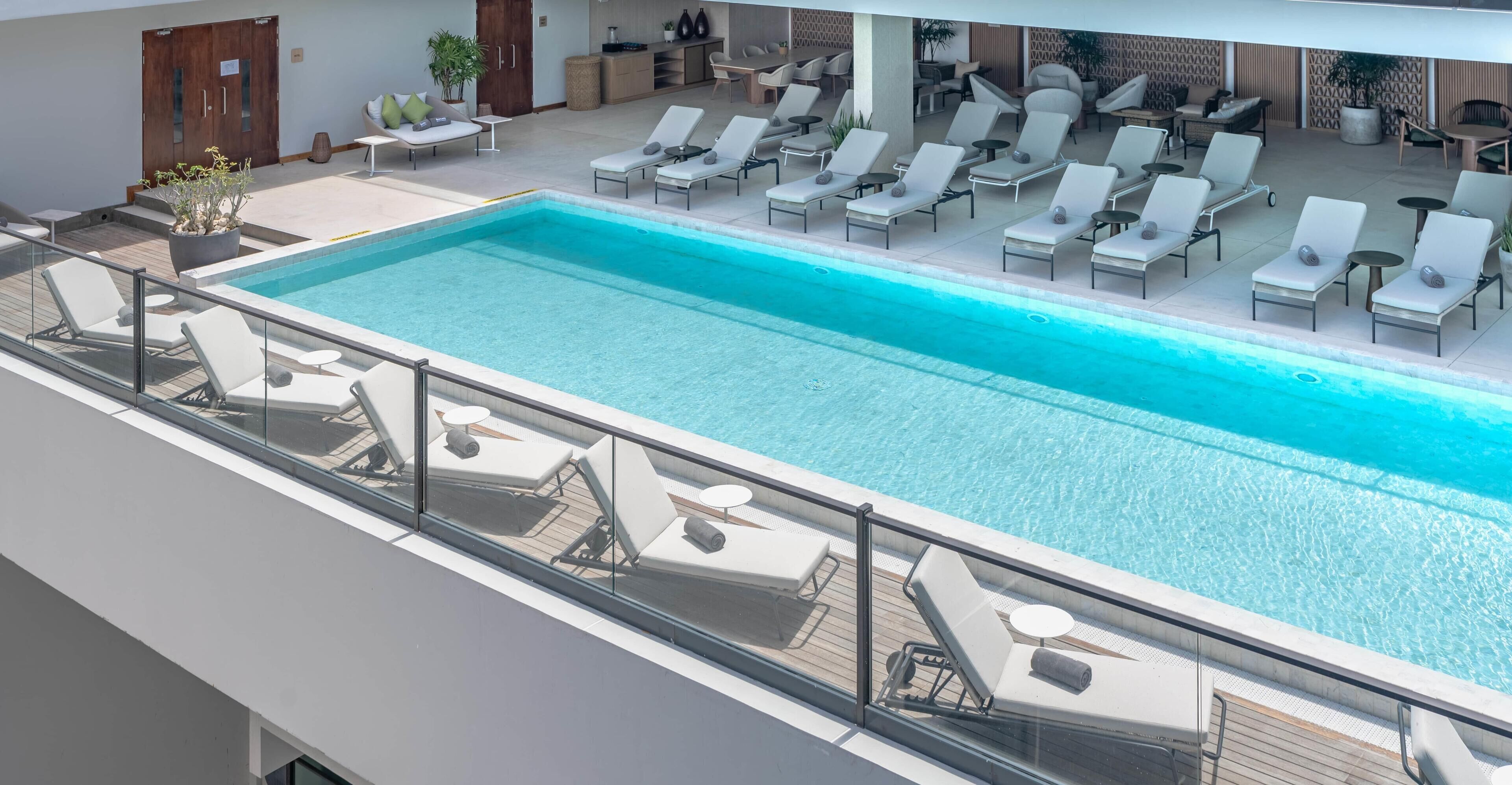 Área de piscina no terraço com espreguiçadeiras e uma pequena mesa, cercada por um deck de madeira e guarda-corpo de vidro.