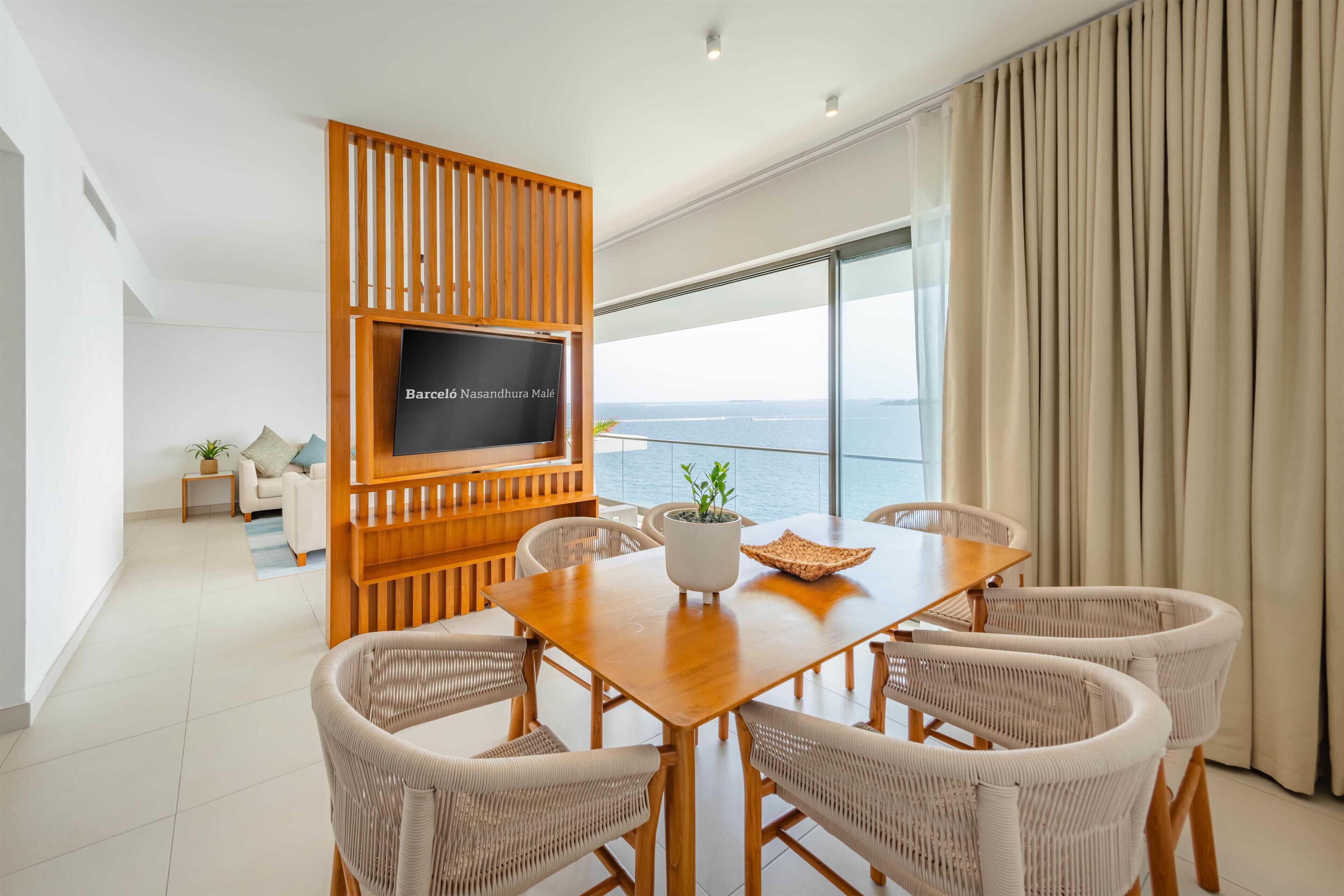 Uma sala de estar moderna com um móvel para TV de madeira, uma mesa de jantar com cadeiras de vime, uma planta em vaso e vista para o mar através de amplas janelas.