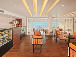 Um café moderno com balcão de madeira, cadeiras laranja e grandes janelas com vista para o mar.