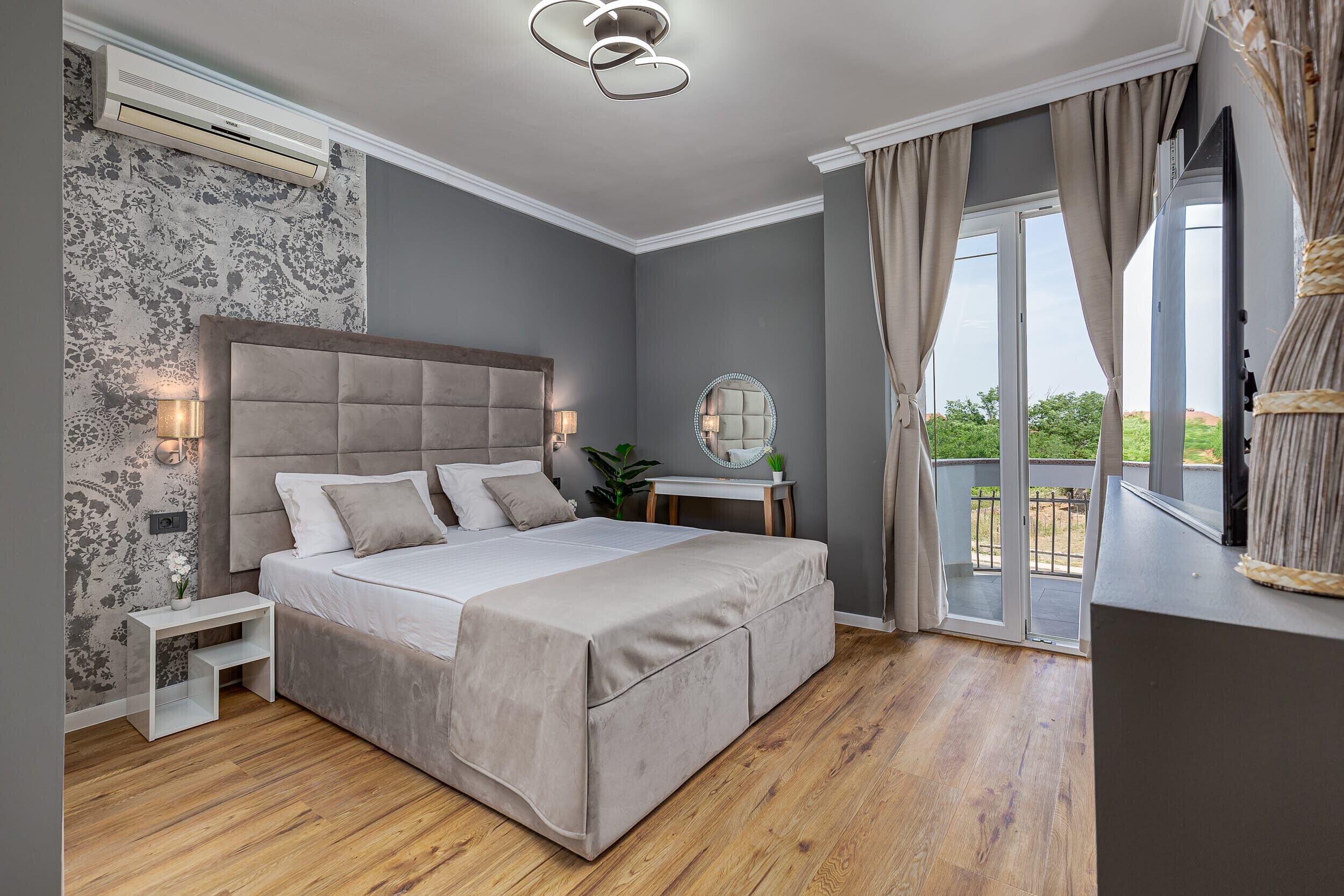 Quarto com cama grande, mesa de cabeceira, espelho e vista para o verde pela janela.
