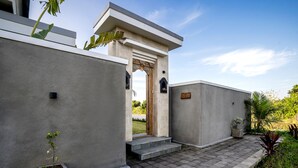 Um edifício moderno com fachada de concreto, porta de madeira e uma pequena área ajardinada com plantas em vasos.