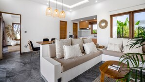 Uma sala moderna com sofá, mesa de centro e luminárias pendentes. Há uma área de jantar com cadeira e mesa, e uma cozinha com armários de madeira.