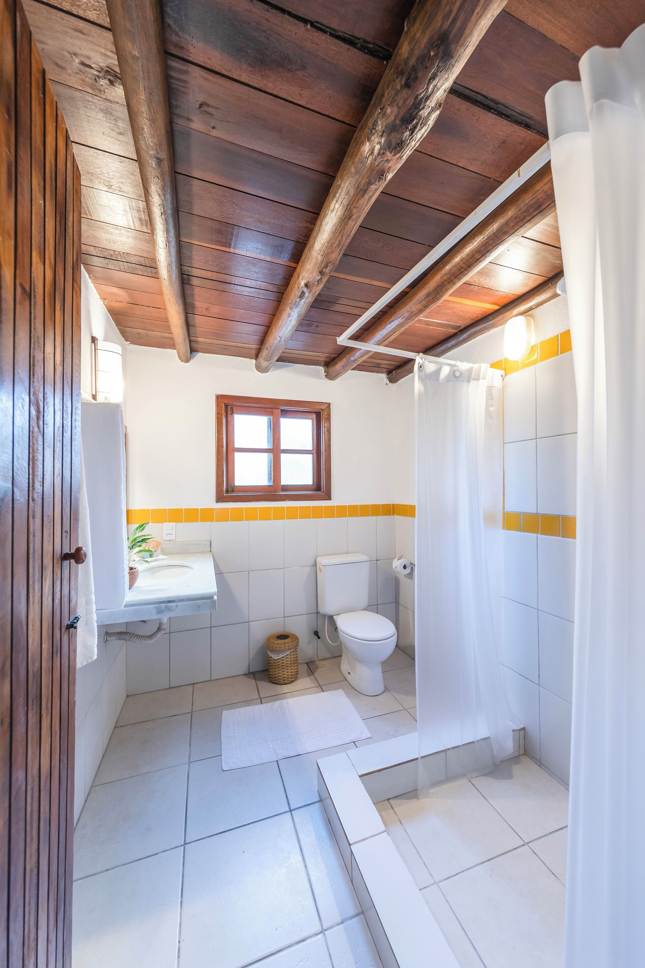 Um banheiro com teto de madeira, azulejos brancos, uma janela, pia, vaso sanitário e cortina de chuveiro.