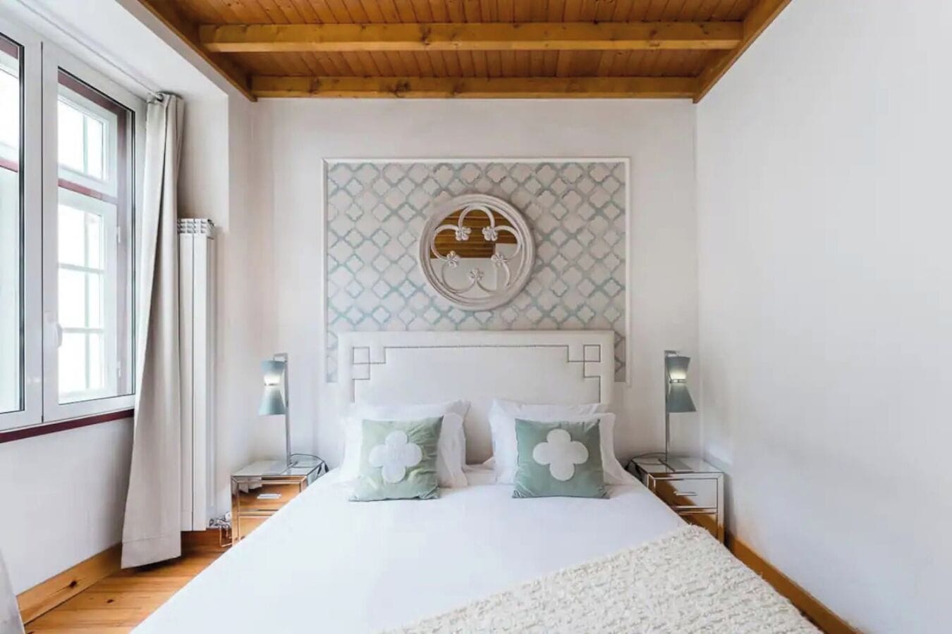 Quarto com teto de madeira, cama com roupa de cama branca e almofadas florais, um detalhe decorativo na parede acima da cama e janela com cortinas.