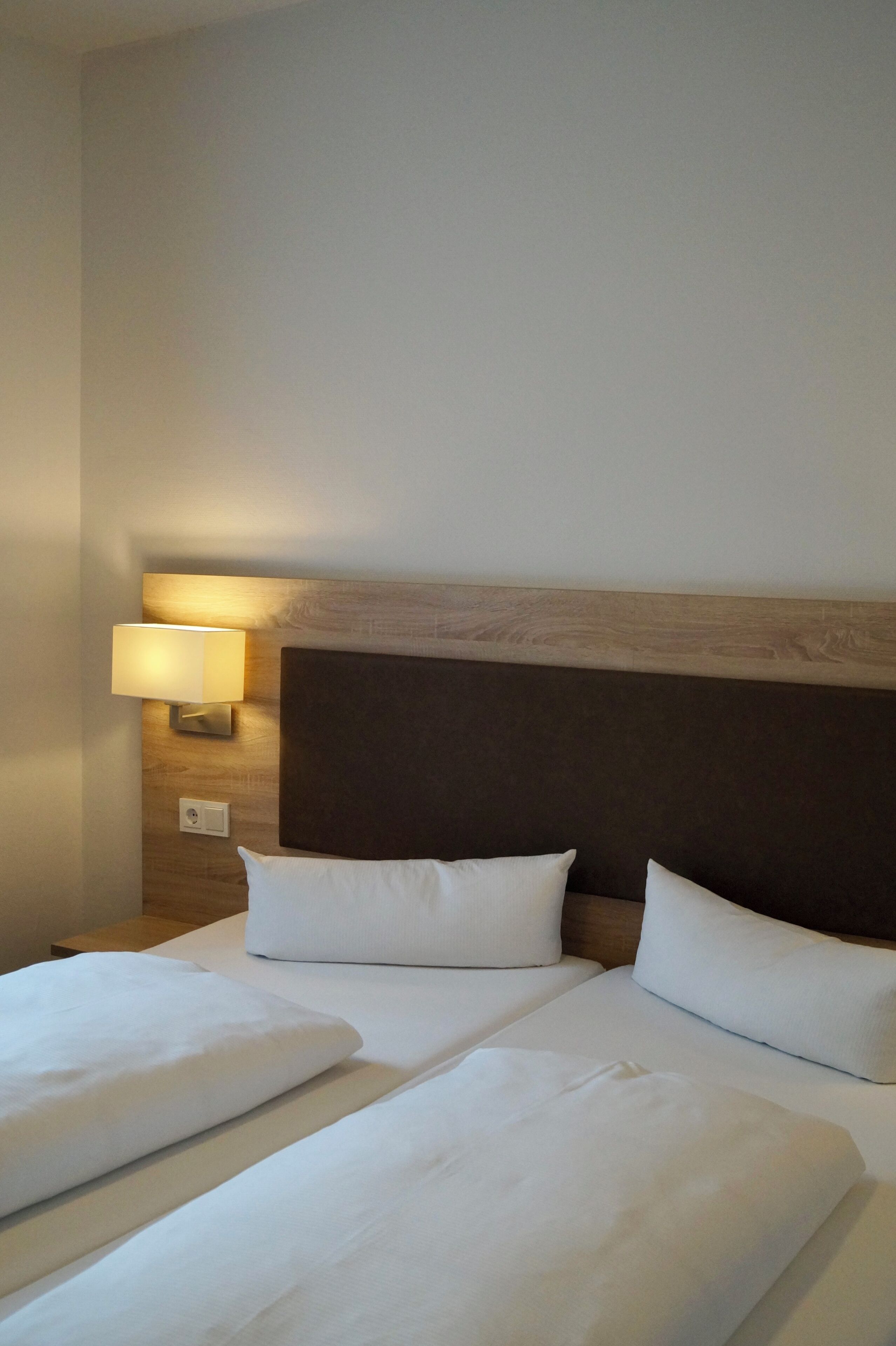 Quarto de hotel com cabeceira de madeira, duas camas com roupa de cama branca e uma luminária fixada na parede.