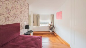 Quarto com uma cama vermelha, uma mesinha com abajur, uma janela com cortinas e um vaso com planta.