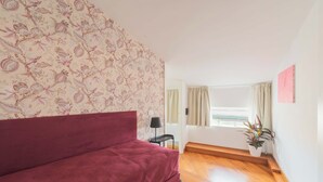 Quarto com sofá vermelho, papel de parede floral, janela com cortinas e planta em vaso.