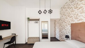 Quarto de hotel com uma cama, escrivaninha com cadeira, televisão e prateleira fixada na parede.