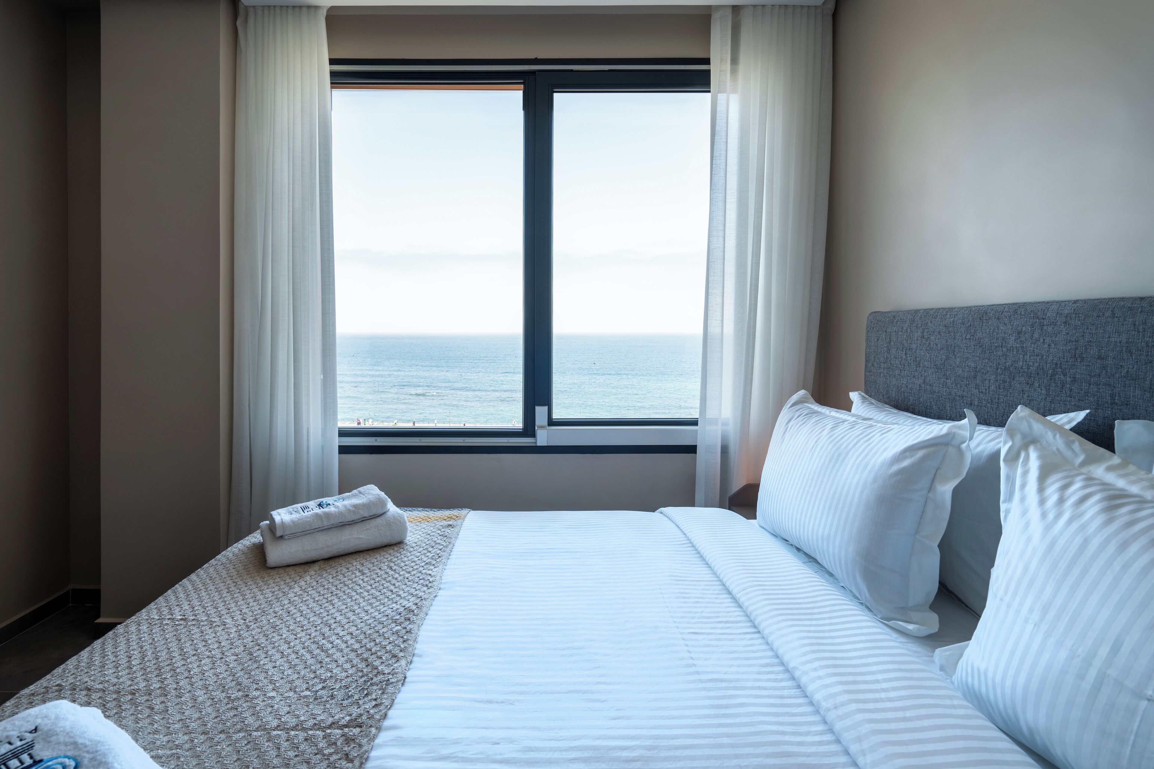 Quarto de hotel com uma cama grande, roupa de cama branca e vista para o mar através de uma janela.