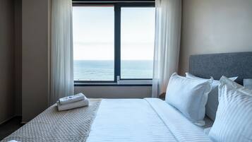 Quarto de hotel com uma cama grande, roupa de cama branca e vista para o mar através de uma janela.