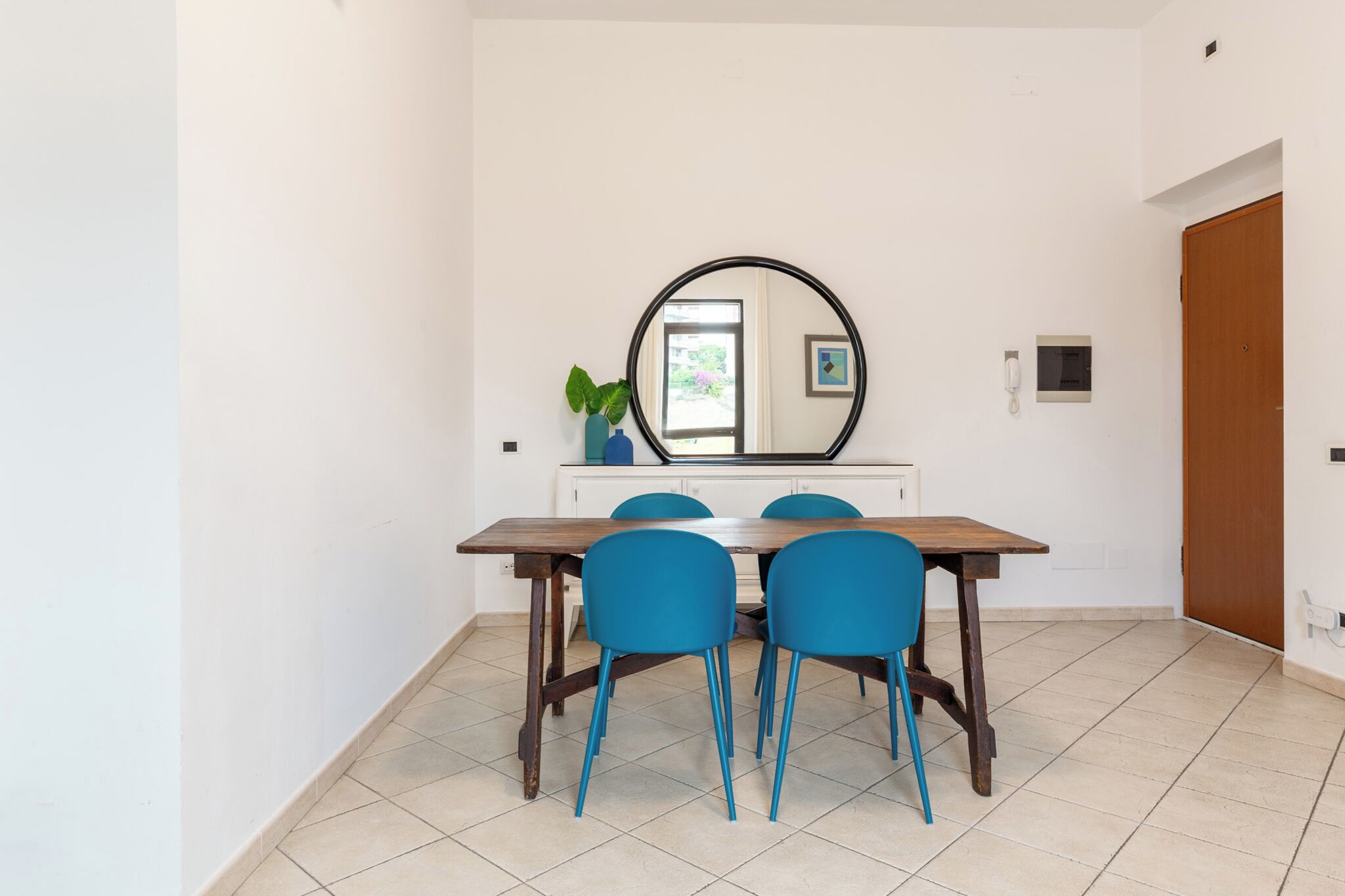 Uma sala de jantar com uma mesa de madeira e cadeiras azuis, uma planta em vaso e um espelho na parede.