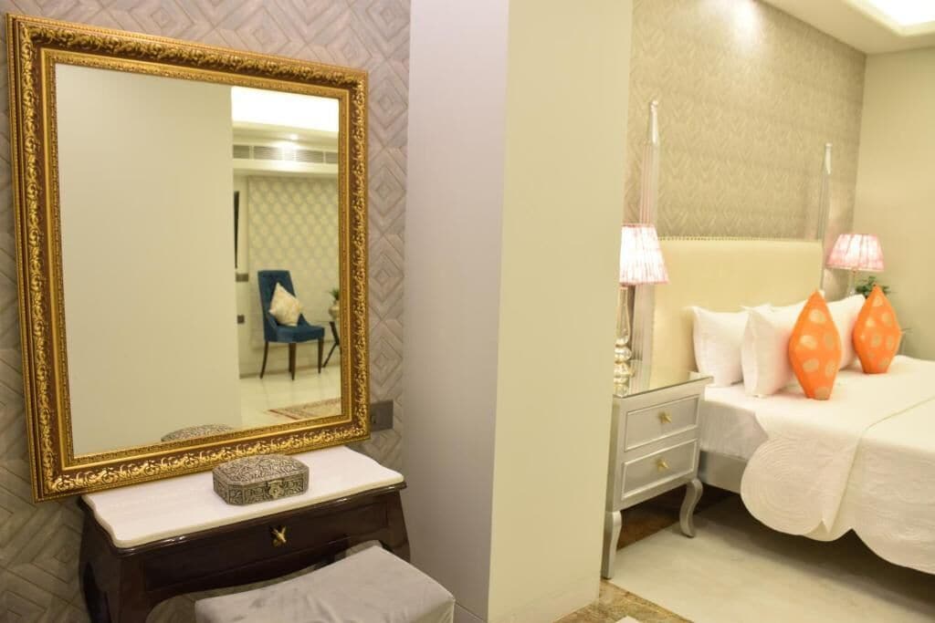 Quarto de hotel com um espelho grande e ornamentado, uma mesa de cabeceira com abajur, uma cama com roupa de cama branca e almofadas laranja, e uma cadeira ao fundo.