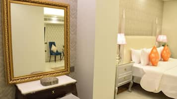 Quarto de hotel com um espelho grande e ornamentado, uma mesa de cabeceira com abajur, uma cama com roupa de cama branca e almofadas laranja, e uma cadeira ao fundo.