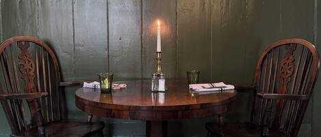 Uma mesa de madeira redonda com duas cadeiras, uma vela acesa, saleiro e pimenteiro, e uma parede verde com um aplique de parede.