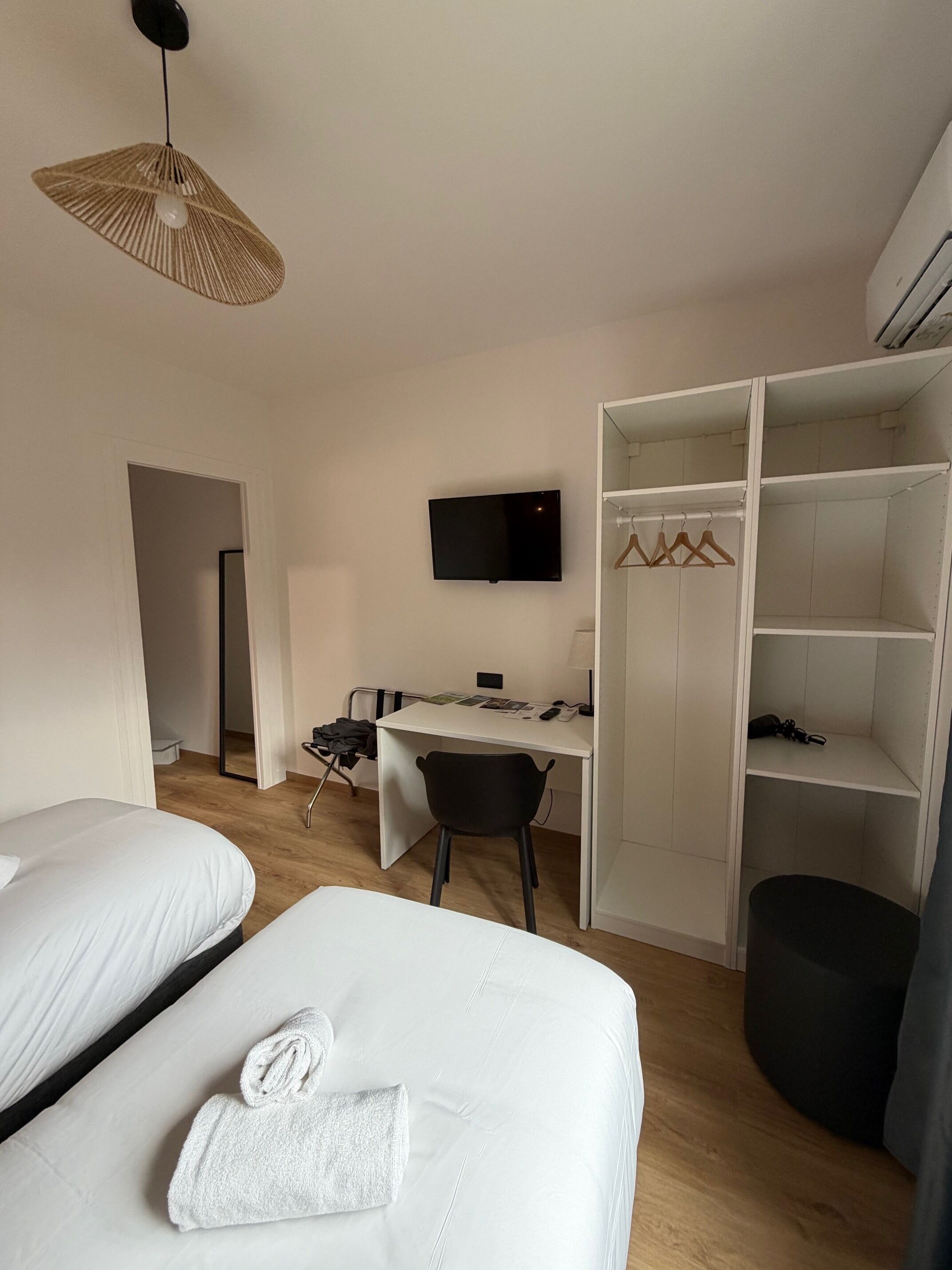 Quarto de hotel com duas camas, uma escrivaninha com cadeira, televisão, guarda-roupa e luminária suspensa.