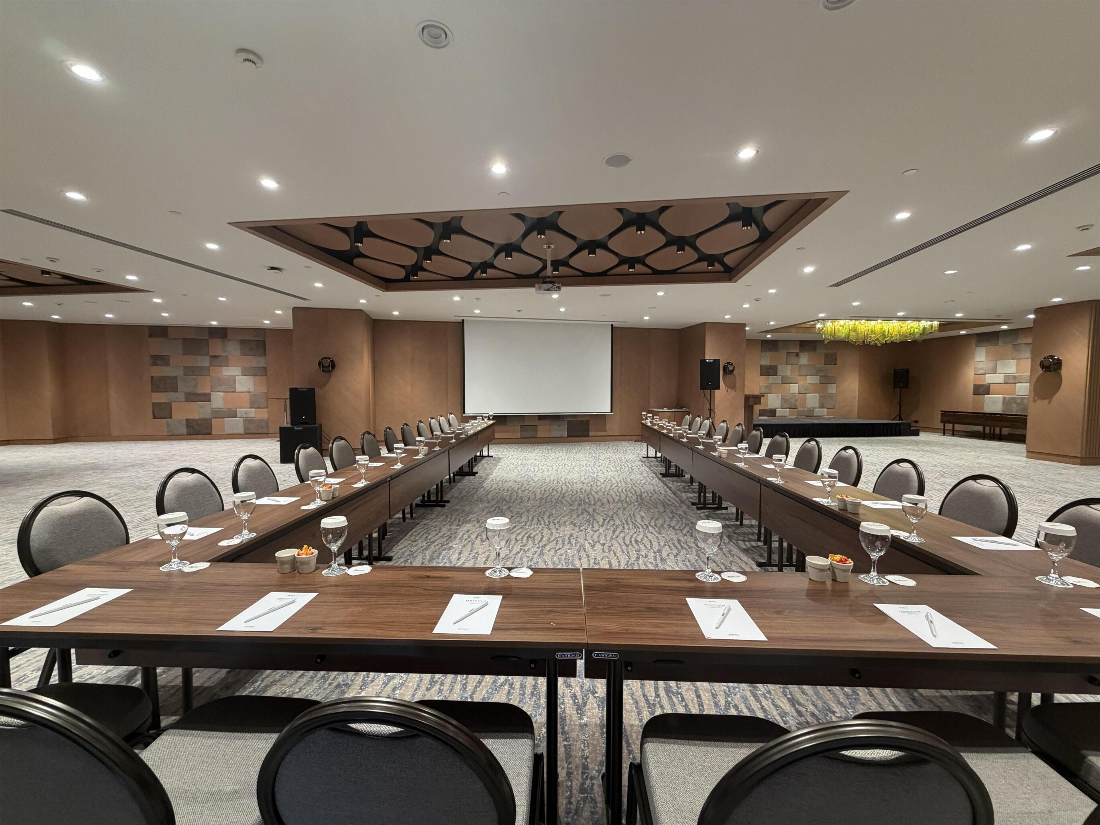 Uma sala de conferências com uma mesa comprida, cadeiras, uma tela de projetor e um palestrante.