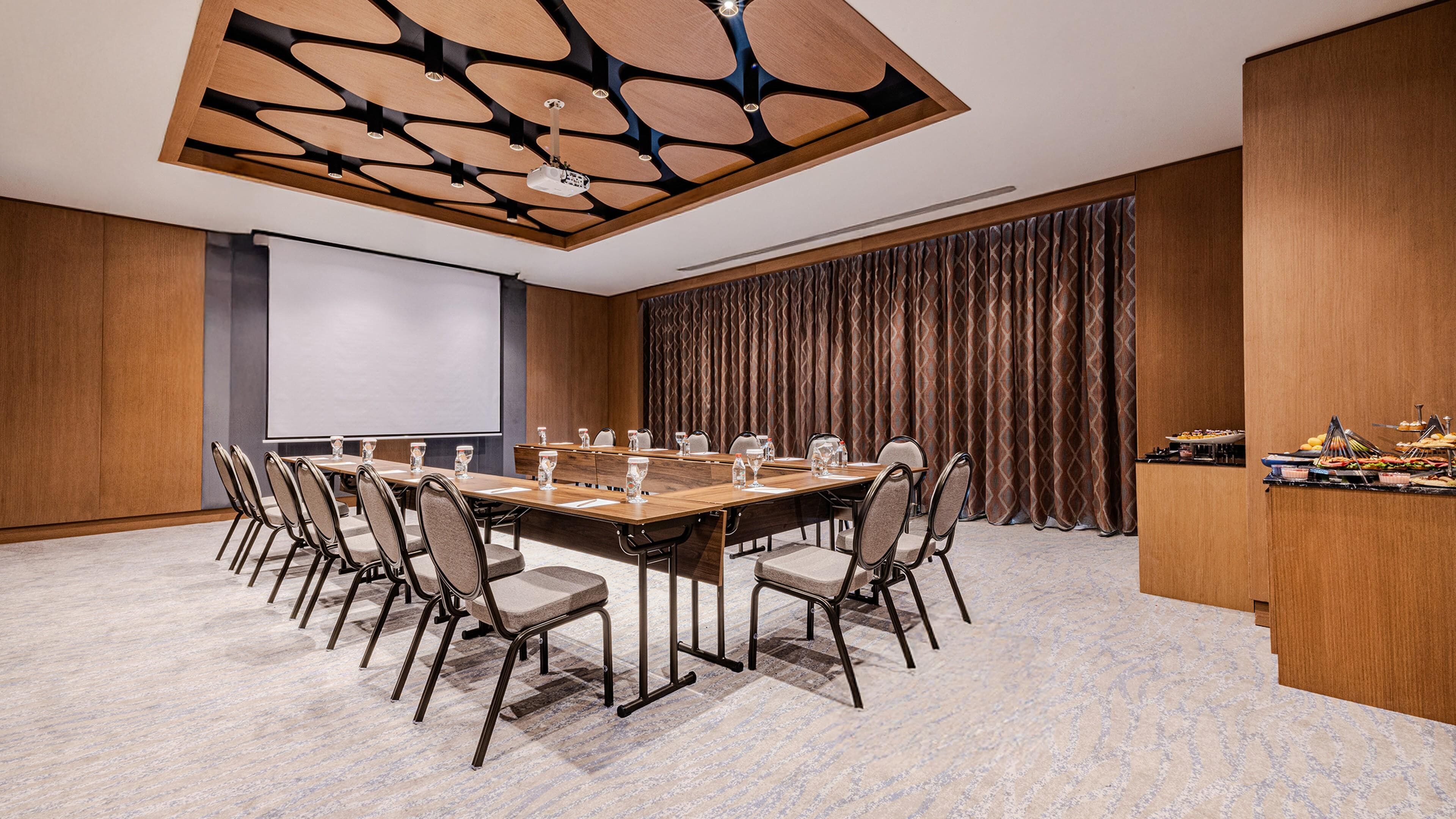 Uma sala de conferências com uma mesa comprida, cadeiras, uma tela de projetor e um espaço para buffet.