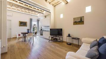 Ampla sala de estar com teto de madeira, mesa de jantar com cadeiras, sofá, televisão e cozinha ao fundo.