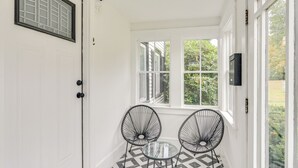 Quarto com porta branca, duas cadeiras de arame pretas, uma mesa de vidro pequena e janela com vista para o verde.