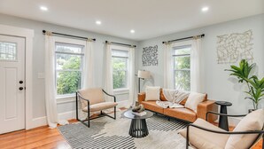 Uma sala de estar moderna com sofá, poltrona e mesa de centro, janelas amplas com cortinas e vista para o verde lá fora.
